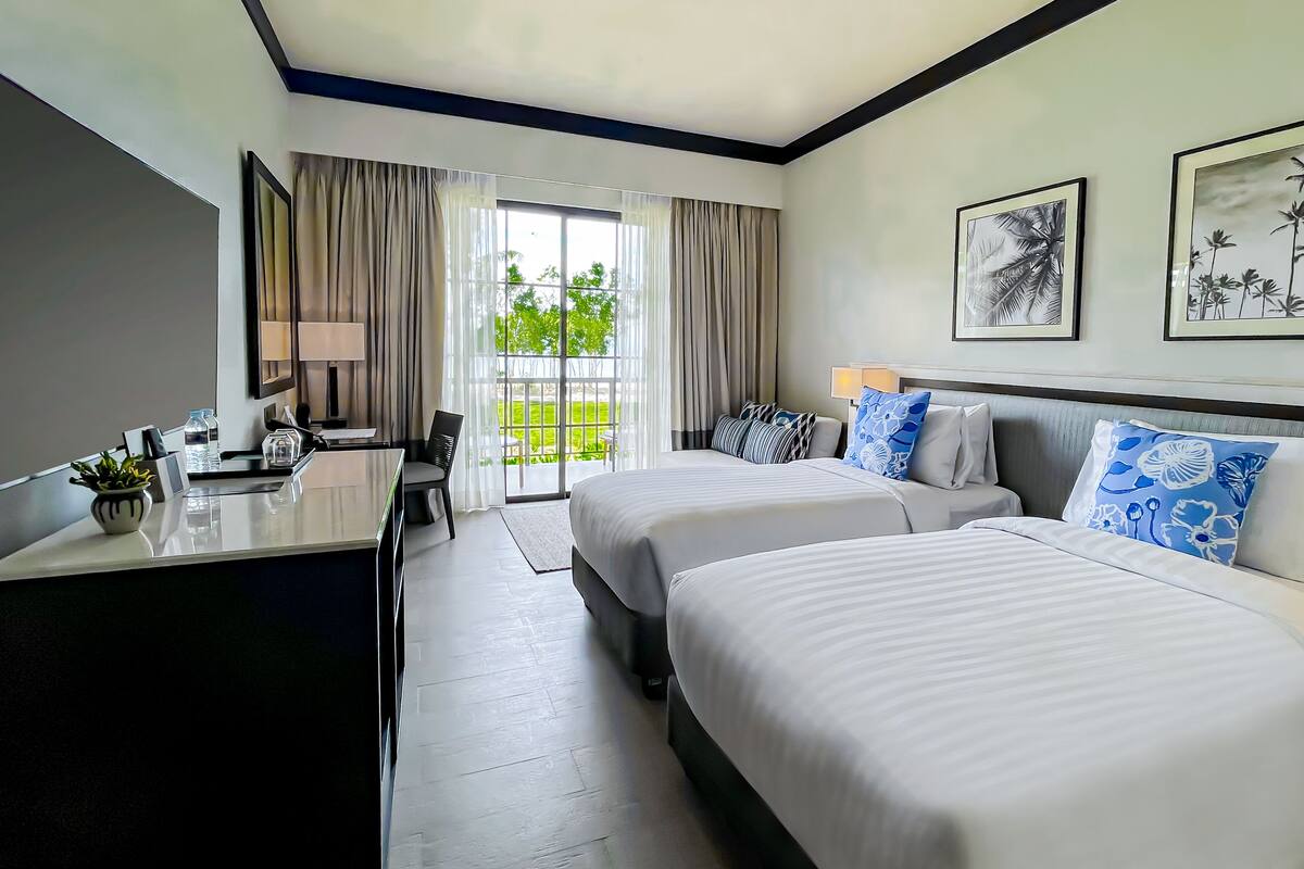 Quarto de hotel com duas camas, uma escrivaninha, uma cadeira, uma televisão e uma janela com cortinas.