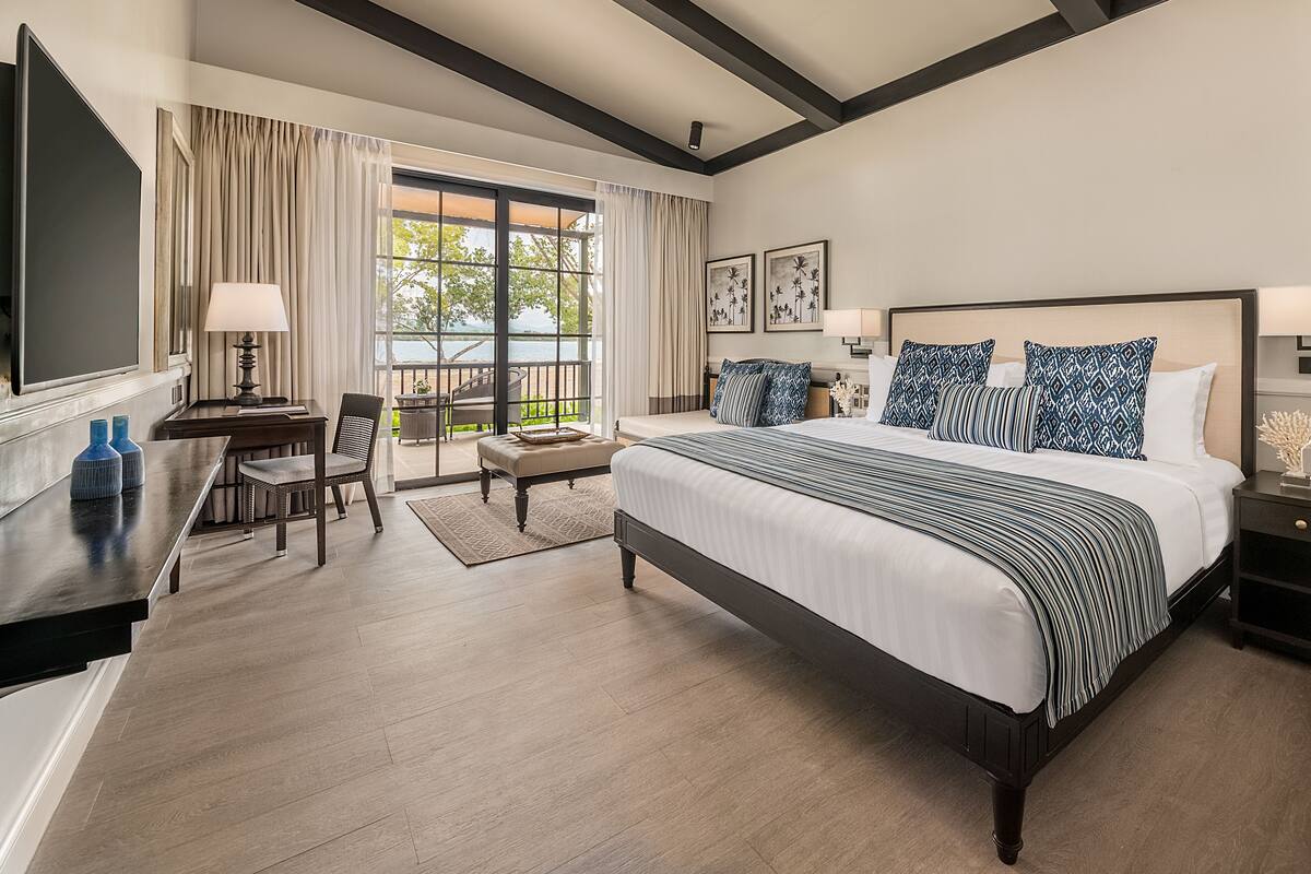 Quarto de hotel com uma cama grande, uma escrivaninha, uma cadeira, varanda com cortinas e um ventilador de teto.