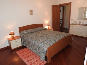 Quarto com cama de madeira, duas mesas de cabeceira com luminárias, cômoda e porta para outro cômodo.