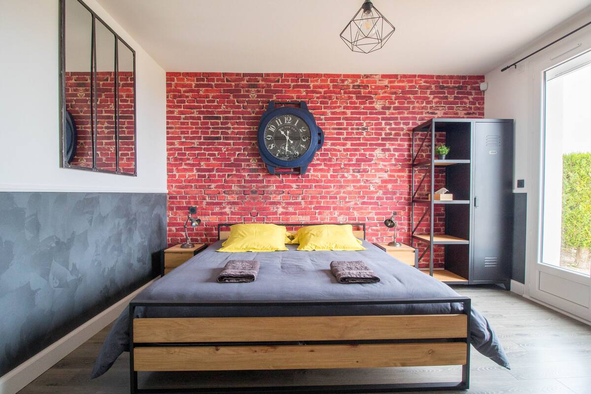 Quarto com parede de tijolos, cama com cabeceira de madeira, um relógio e um guarda-roupa.