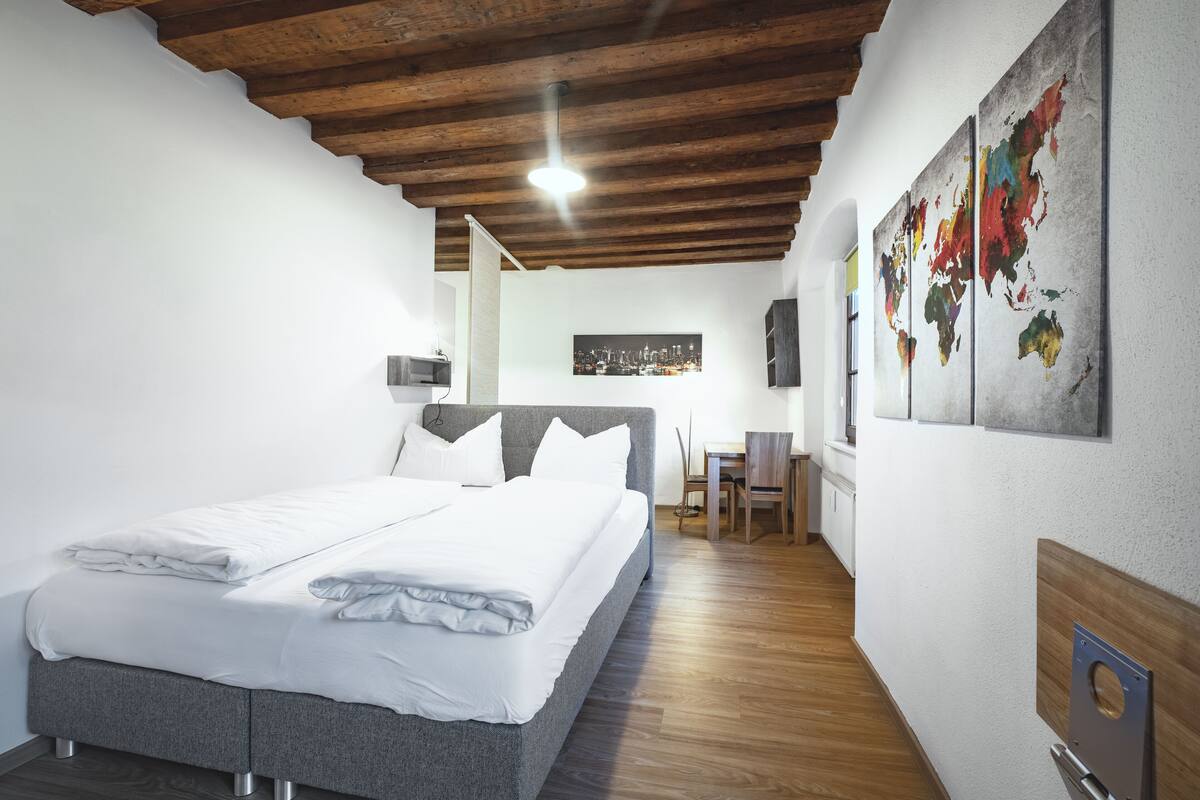Um quarto moderno com uma cama cinzenta, vigas de madeira no teto, uma obra de arte com paisagem urbana e uma área de refeições com mesa e cadeiras.