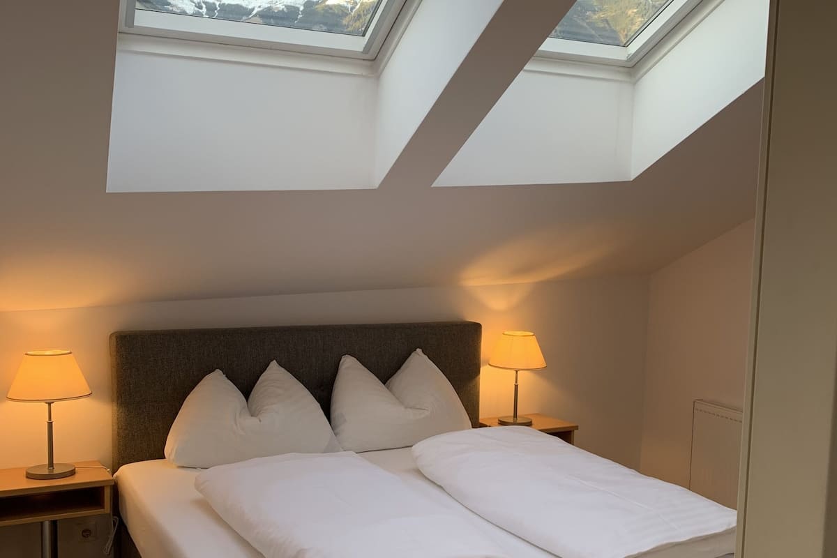 Quarto com cama, duas luminárias de cabeceira, mesa de cabeceira, piso de madeira e janelas no teto com vista para montanhas.