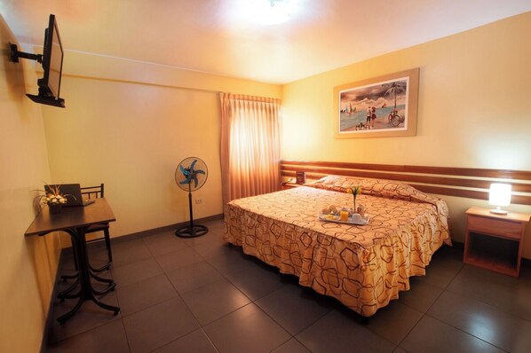 Quarto de hotel com cama, escrivaninha, cadeira, ventilador, mesa de cabeceira e televisão fixada na parede.