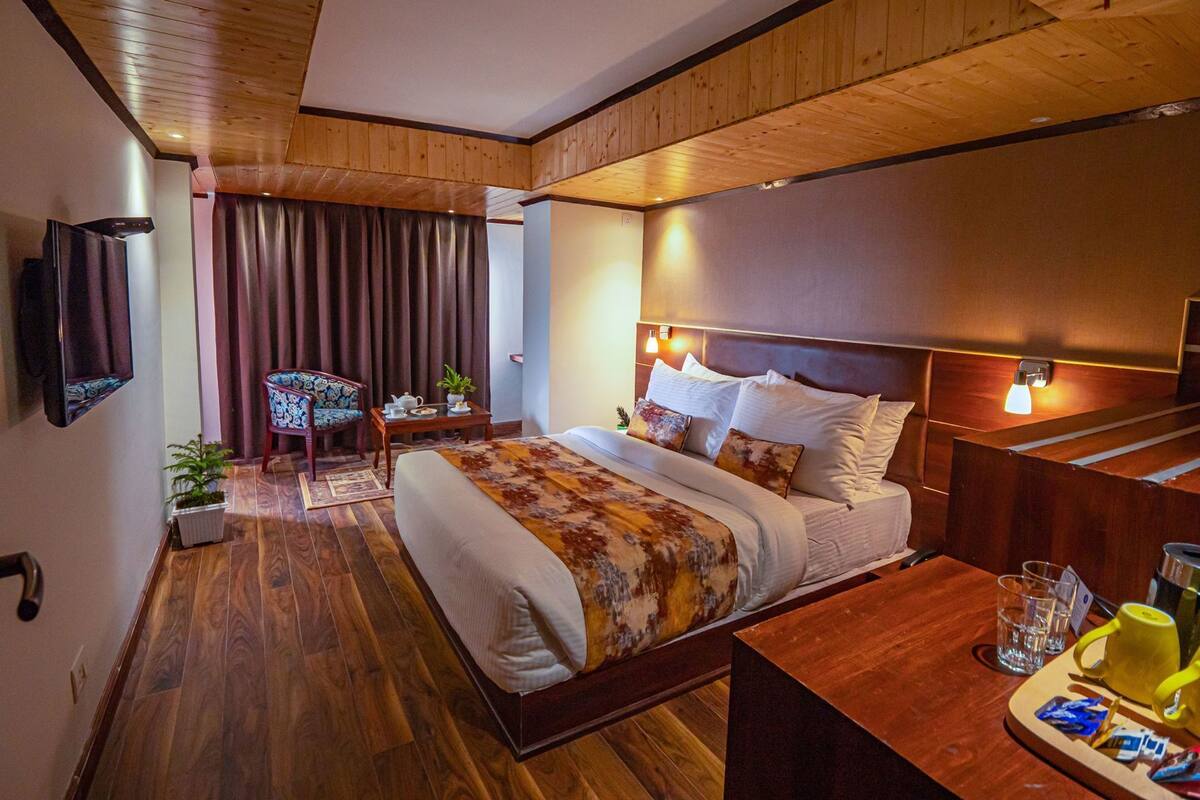 Quarto de hotel com teto de madeira, cama com colcha estampada, mesa de cabeceira com luminária, uma cadeira e televisão fixada na parede.