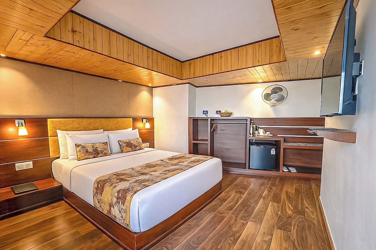 Quarto de hotel com teto de madeira, cama com colcha estampada, pequena mesa de escritório com um computador e um ventilador fixado na parede.
