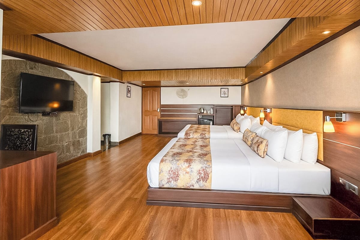 Quarto de hotel com cama grande, piso de madeira, lareira de pedra, televisão e cozinha compacta.