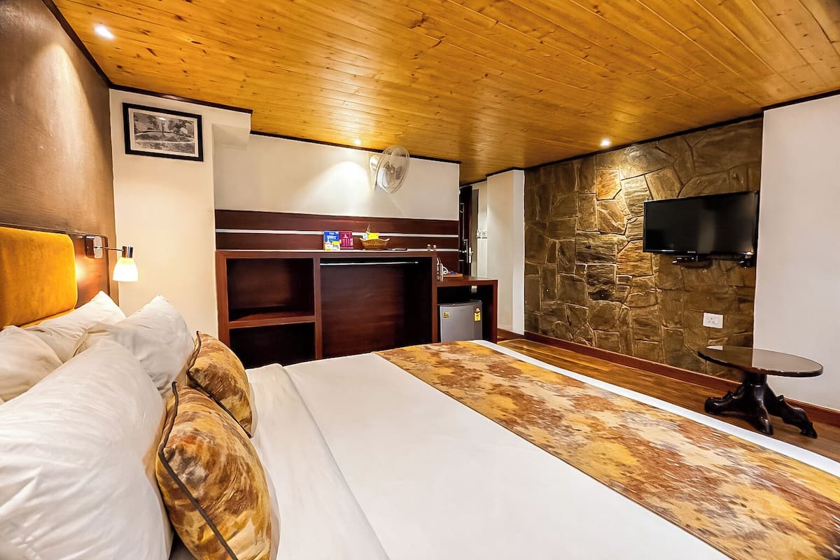 Quarto de hotel com teto de madeira, cama com roupa de cama branca, parede de pedra, televisão e uma pequena mesa.