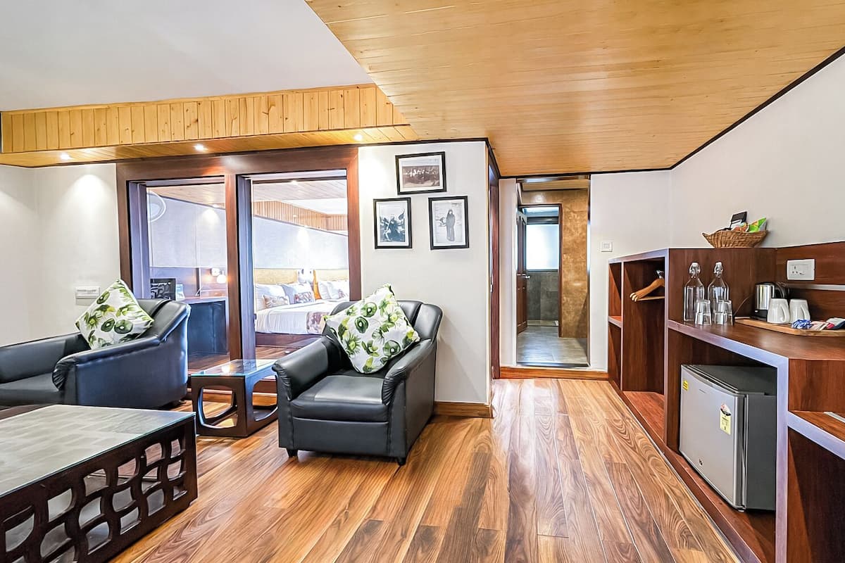 Quarto de hotel moderno com teto de madeira, área de estar com duas cadeiras e uma mesa, cozinha compacta e vista para uma varanda através de uma porta de vidro.