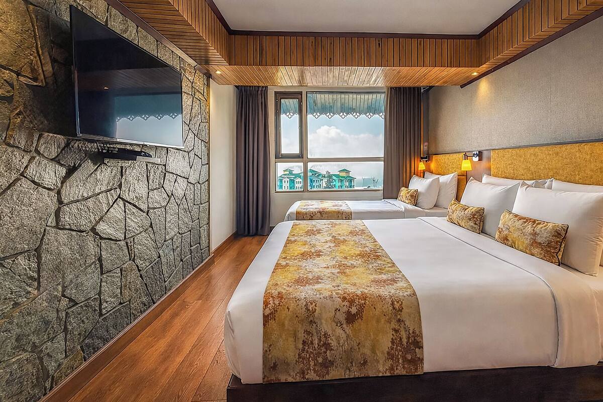 Quarto de hotel com parede de pedra, televisão fixada, cama grande com roupa de cama branca, cabeceira com padrão decorativo e janela com cortinas.