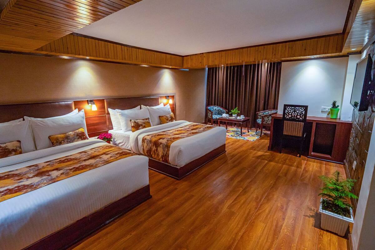 Quarto de hotel com duas camas, uma mesa, uma cadeira, uma mesinha e um vaso de planta.