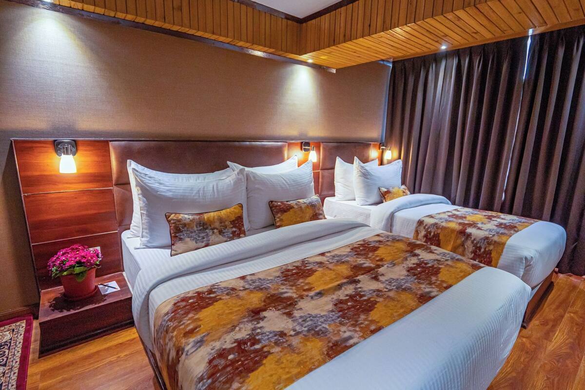 Quarto de hotel com duas camas, cabeceira de madeira, luminárias fixadas na parede e um vaso com planta.