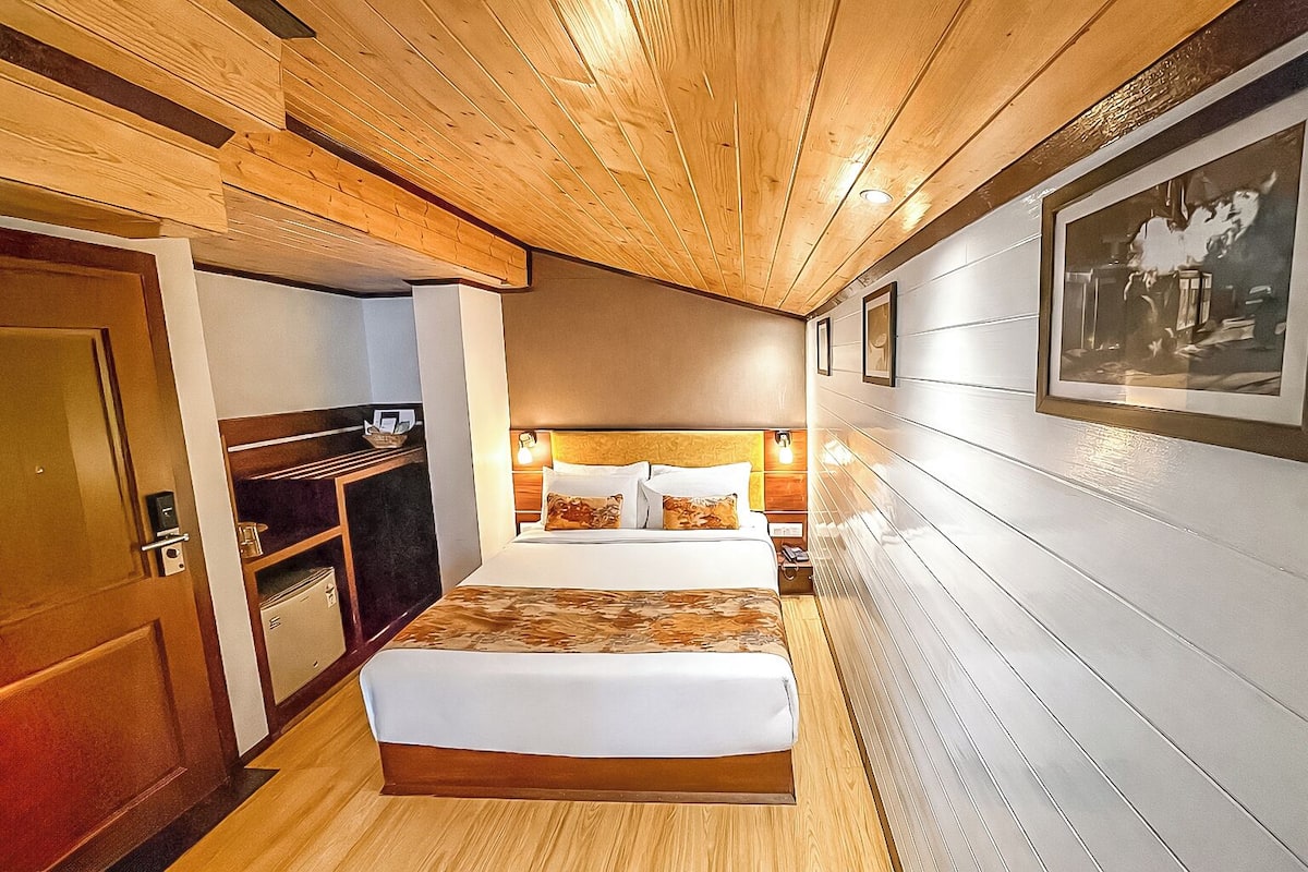 Quarto de hotel com teto de madeira, cama com colcha estampada, mesa de cabeceira, televisão presa à parede e um quadro emoldurado.
