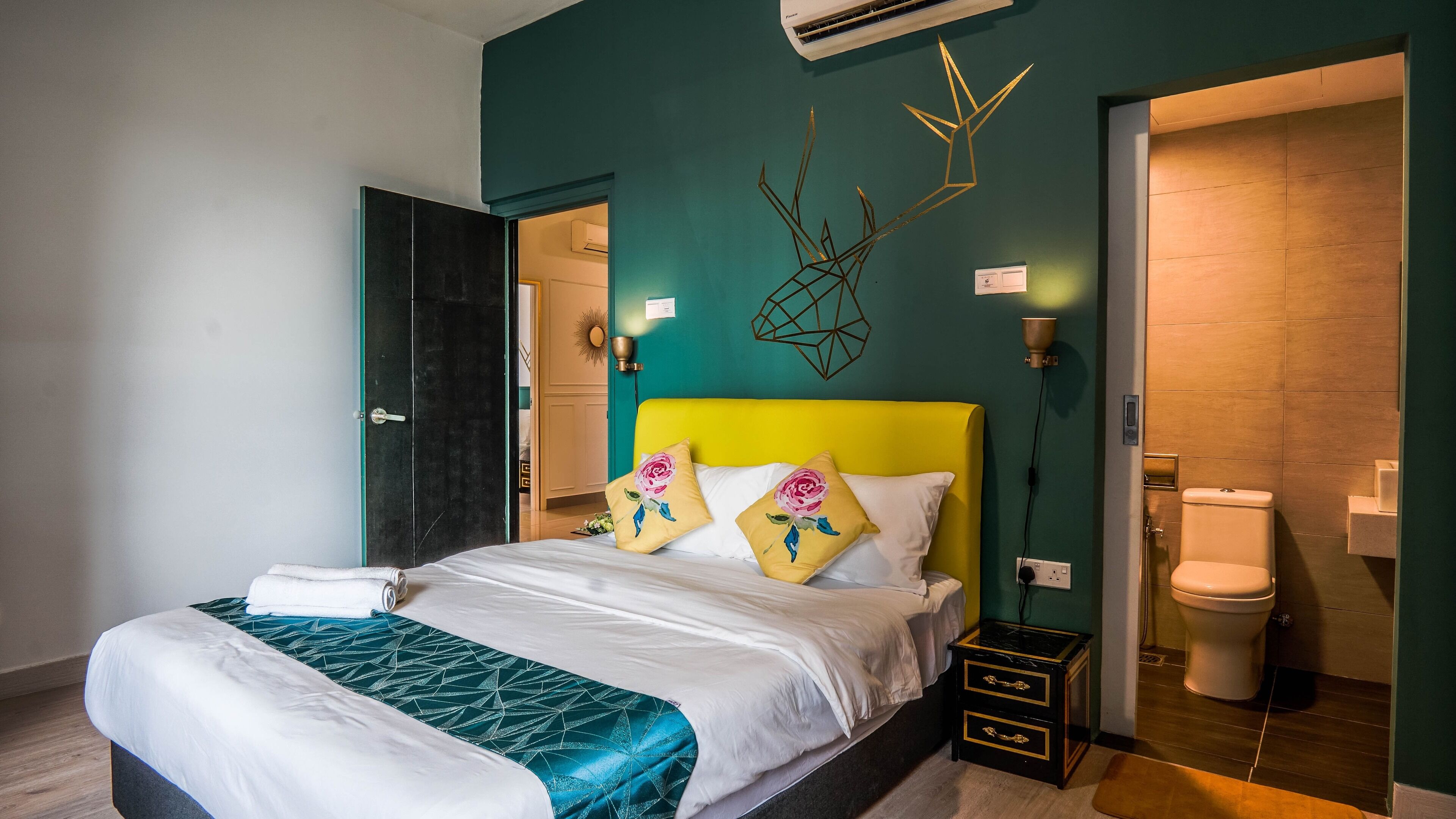 Quarto de hotel moderno com uma cama grande, paredes verde-água e cabeceira amarela, e um banheiro com vaso sanitário e pia.