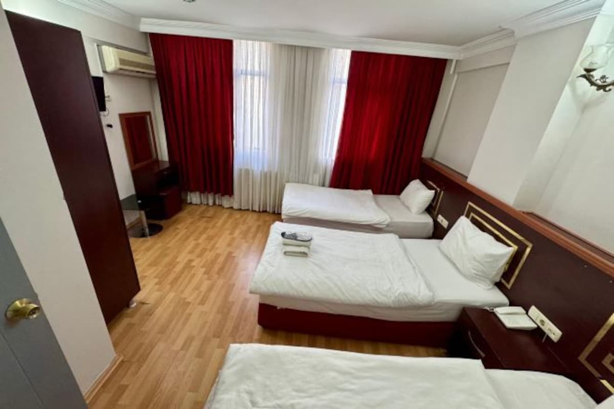 Quarto de hotel com duas camas, piso de madeira, escrivaninha e janela com cortinas vermelhas.