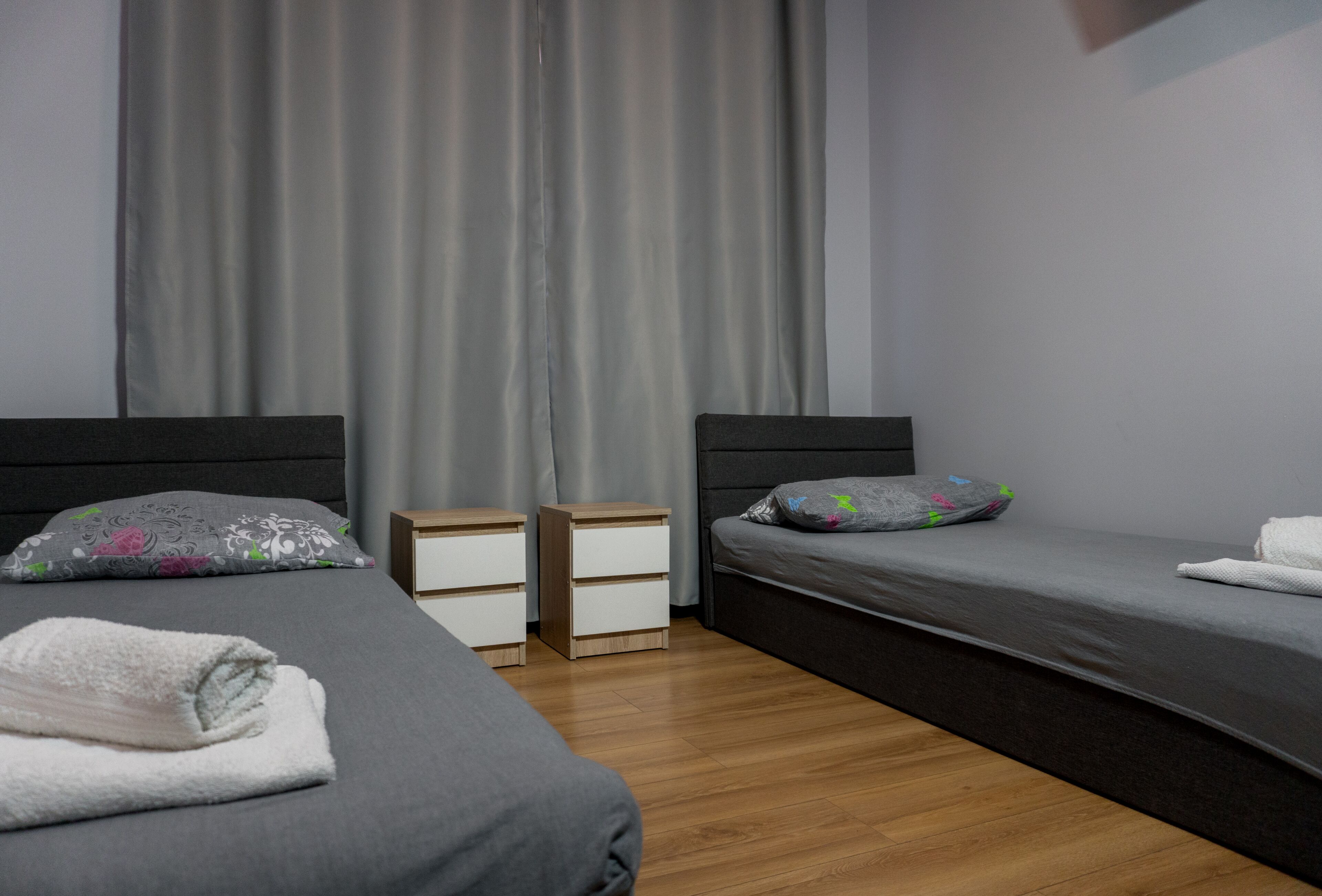 Quarto com duas camas, mesas de cabeceira de madeira e cortina cinza.