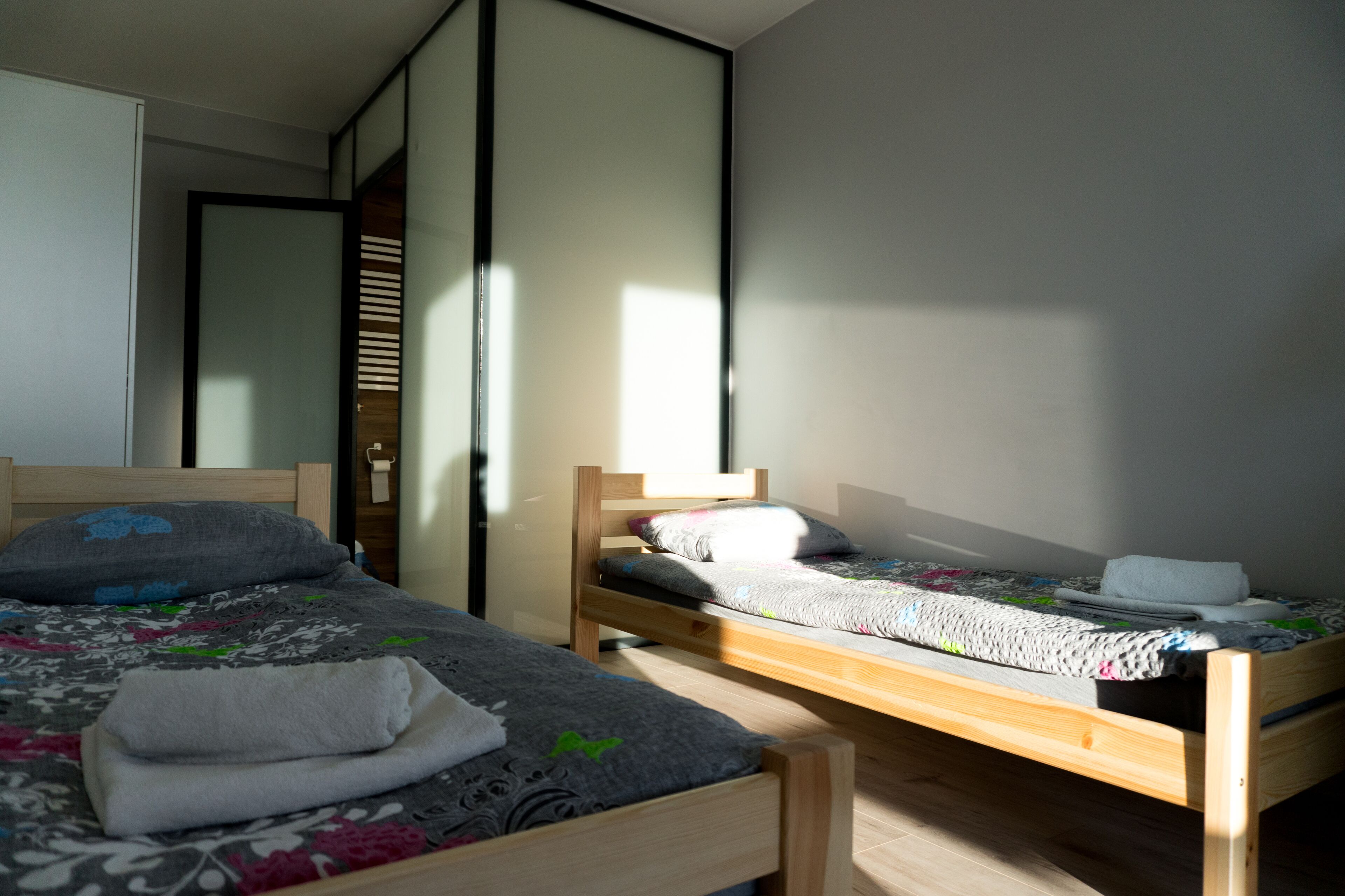 Um quarto com duas camas de madeira, uma com colcha cinza estampada e outra com colcha branca.