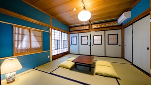 Um quarto japonês tradicional com piso de tatame, uma mesa de madeira baixa, almofadas, portas deslizantes e um ventilador de teto.
