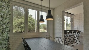 Uma sala de jantar com mesa e cadeiras de madeira, janelas amplas e luminárias pendentes.