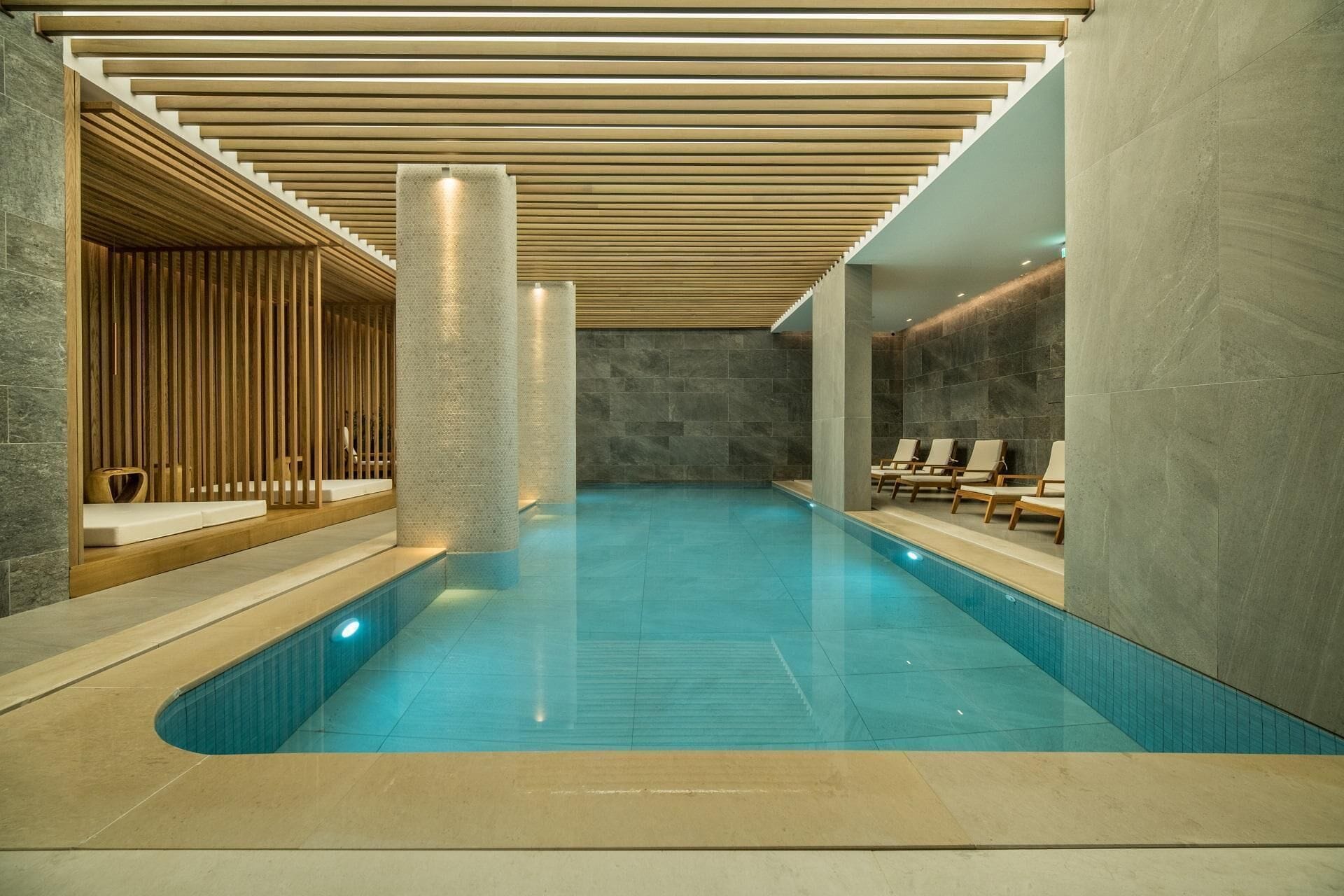 Piscina interior moderna com superfície de água azul cristalina, teto de ripas de madeira e paredes de pedra.