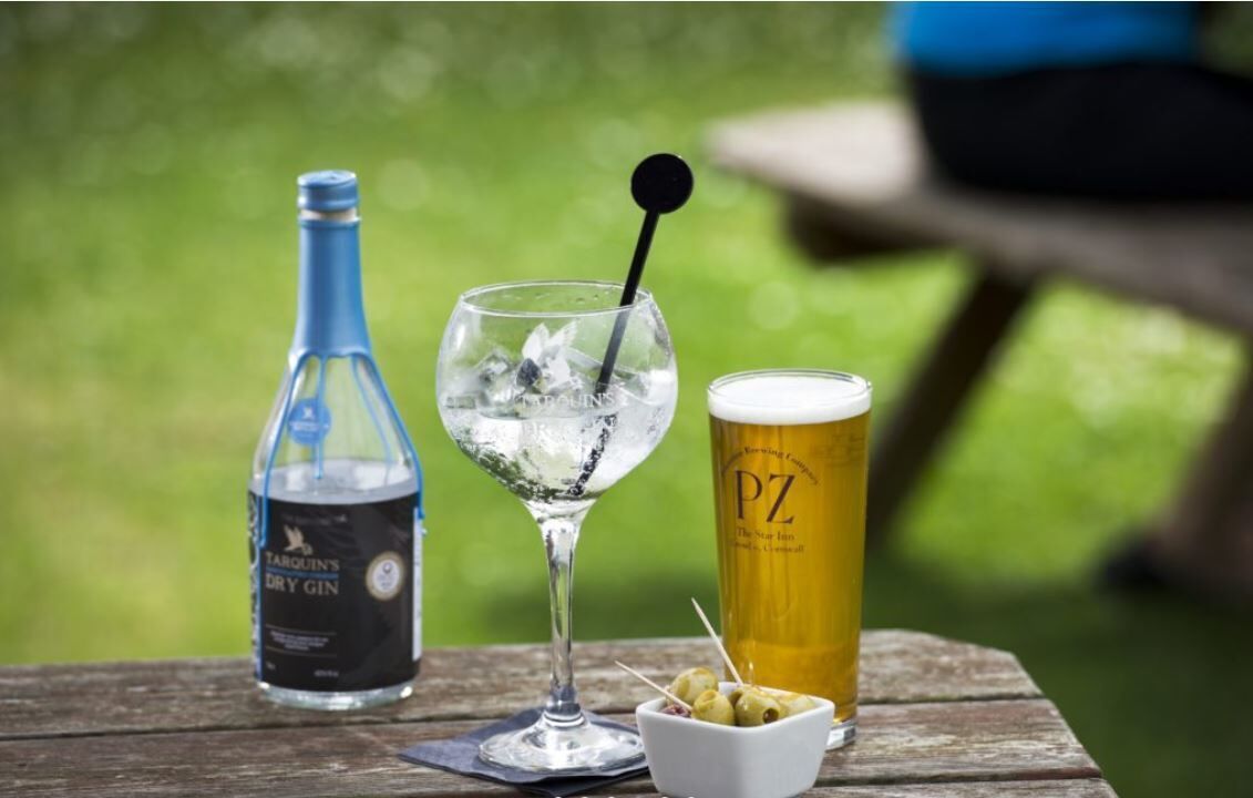 Uma garrafa de Gin Seco Tarquin, um copo com gelo e um mexedor de coquetel, e um copo alto de cerveja sobre uma mesa de madeira.