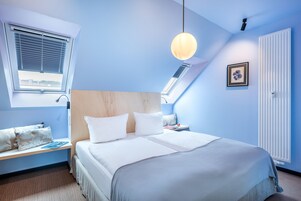 Quarto com claraboia, cama com roupa de cama branca, mesa de cabeceira com abajur e um quadro na parede.