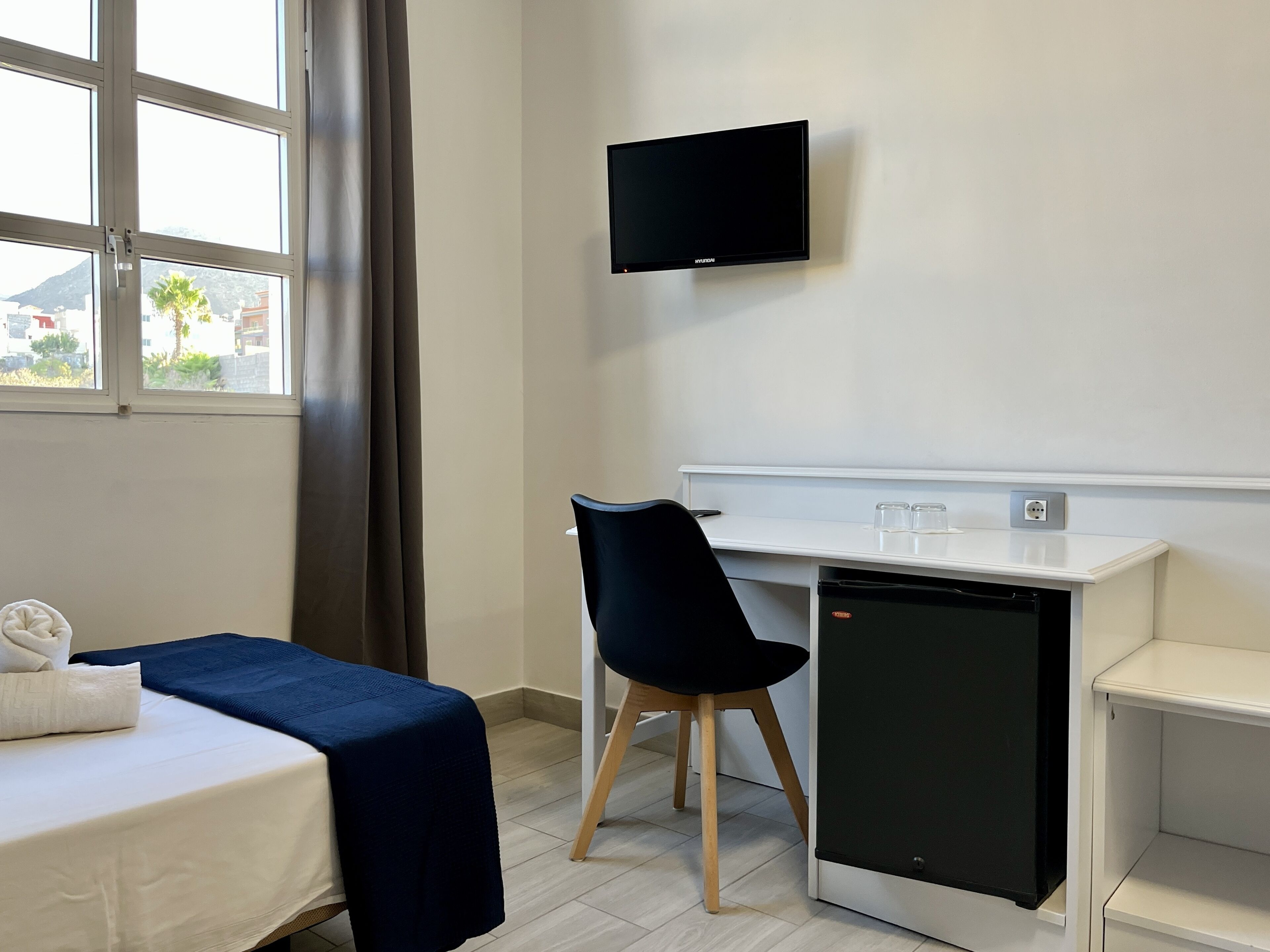 Quarto de hotel com cama, escrivaninha com computador, televisão e pequeno frigorífico.
