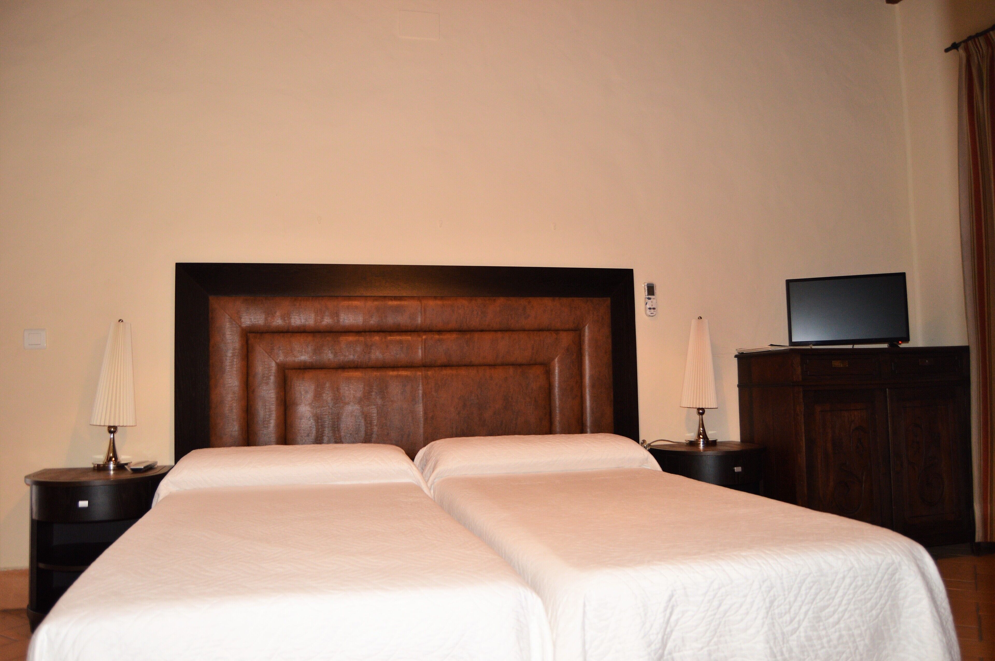 Quarto de hotel com duas camas, cabeceira de madeira, televisão sobre um móvel e duas luminárias de cabeceira.