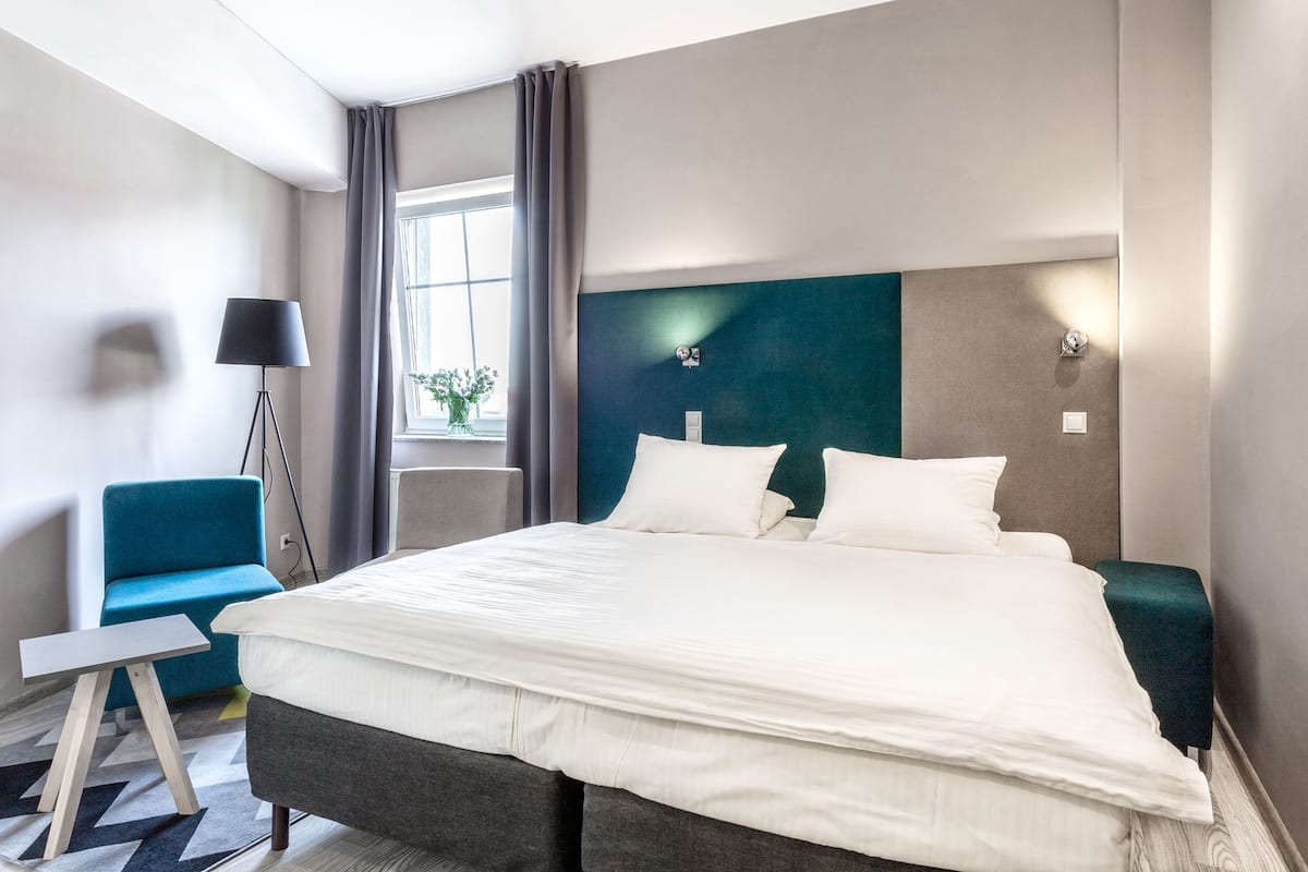 Quarto de hotel moderno com uma cama grande, uma escrivaninha, uma cadeira e uma janela com cortinas.