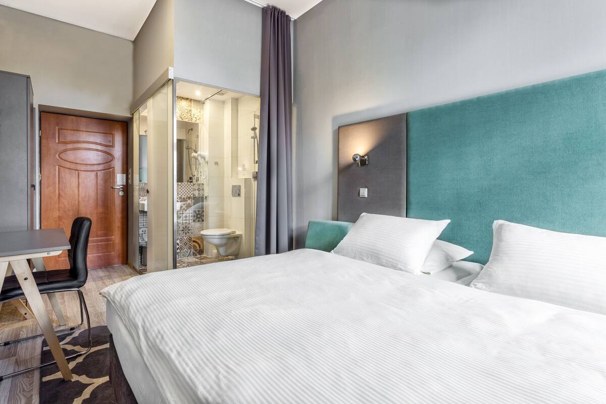 Quarto de hotel com cama, escrivaninha, cadeira e banheiro visível através de uma porta de vidro.
