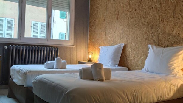 Quarto de hotel com duas camas, uma parede de destaque em madeira, uma janela com persianas e um radiador.