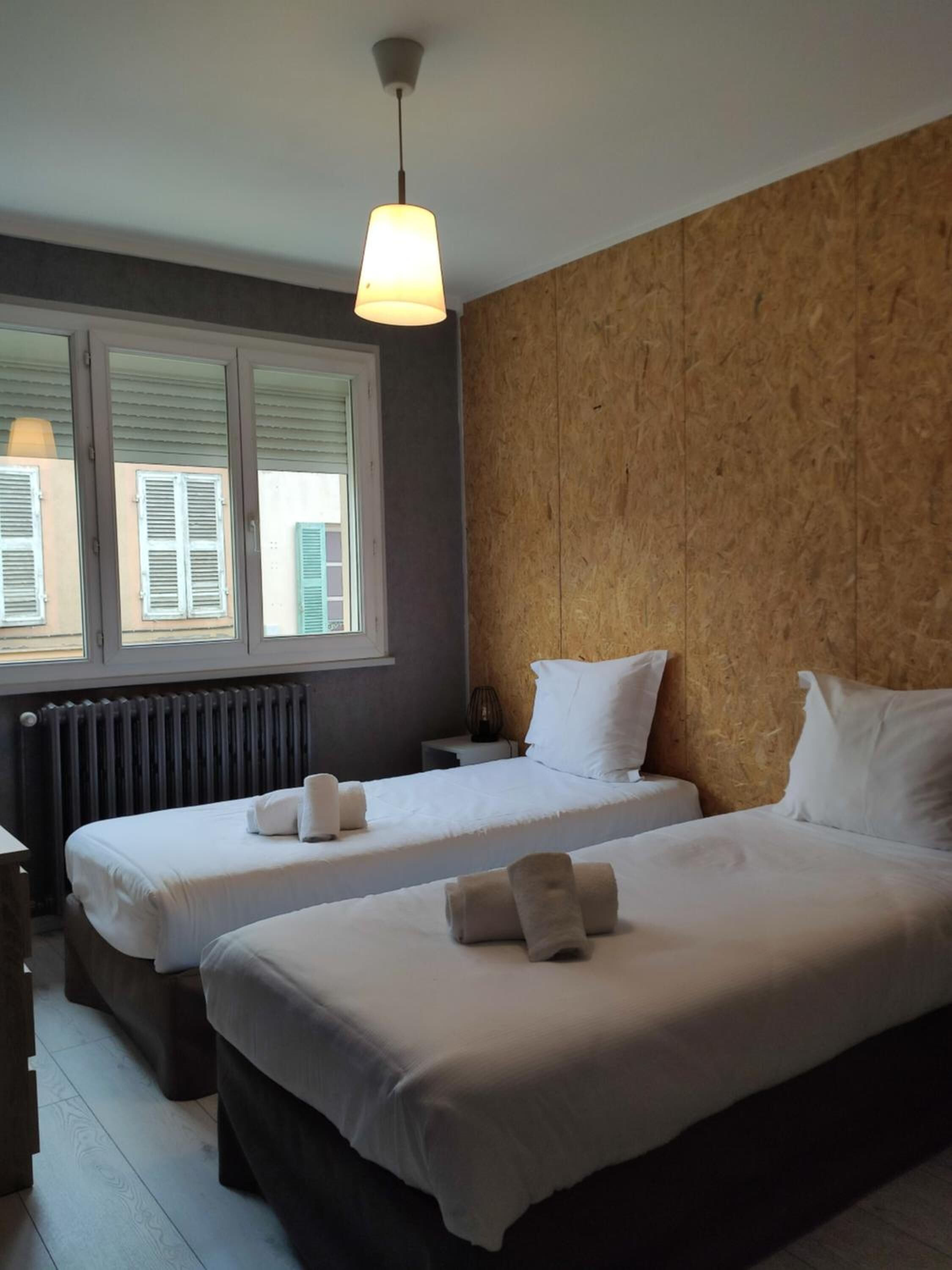 Quarto de hotel com duas camas, cabeceira de madeira, janela com persianas, radiador e luminária pendente.