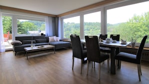 Uma sala de estar moderna com sofá, mesa de jantar e cadeiras, grandes janelas com vista para uma floresta.