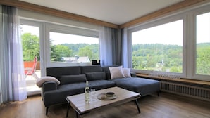 Uma sala de estar moderna com um sofá modular cinza, uma mesa de centro de madeira e grandes janelas com vista para uma área arborizada.