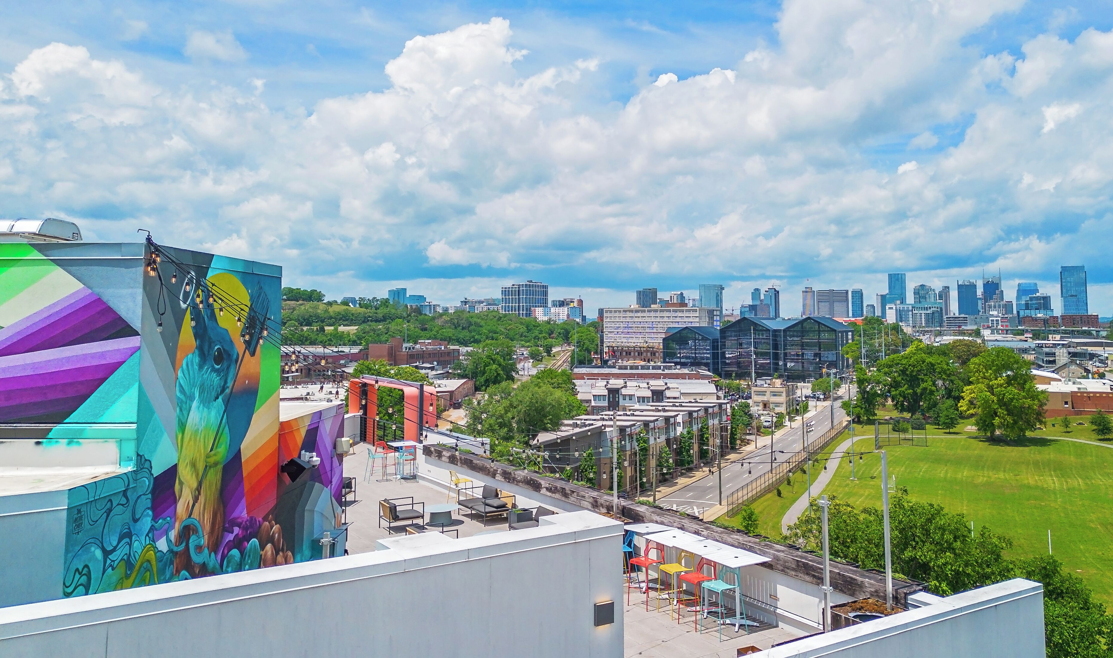 Paisagem urbana com um edifício de destaque adornado por um mural colorido, um parque verde e um céu claro.