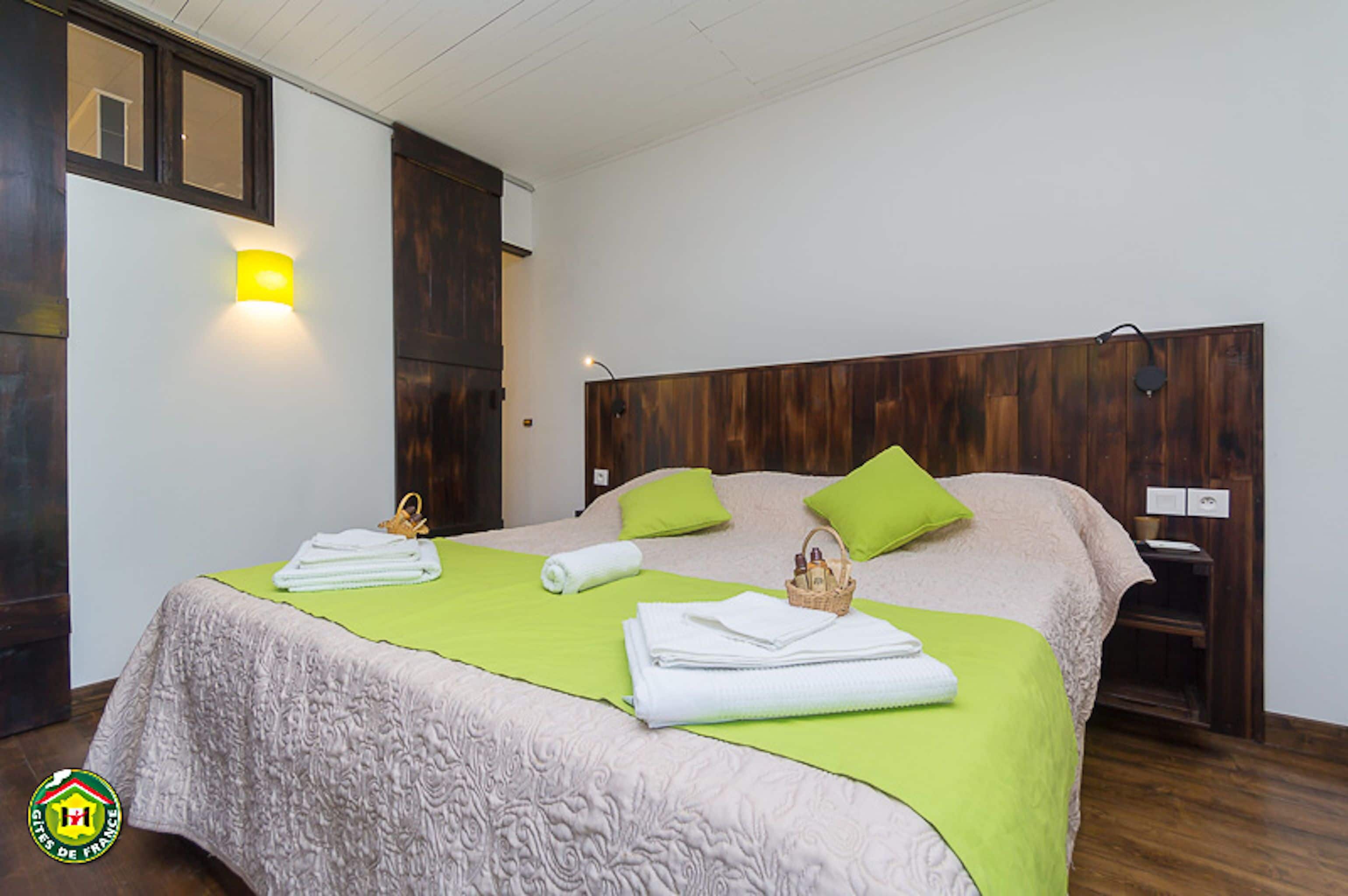 Quarto de hotel com cama grande, roupa de cama verde e cabeceira de madeira. Há toalhas e uma cesta na cama.