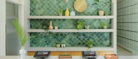 Uma área de recepção com parede estampada em verde, prateleiras com plantas, um objeto decorativo redondo e dois dispositivos digitais no balcão.