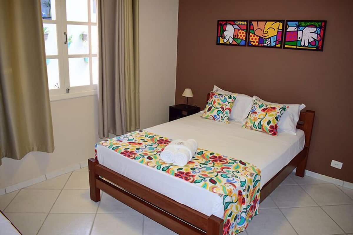 Uma cama bem arrumada com um edredom floral colorido e duas almofadas, uma mesa de cabeceira com abajur e uma janela com cortinas.