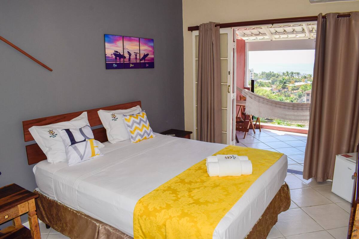 Cama bem arrumada com colcha amarela, uma rede e vista para o oceano através de cortinas abertas.
