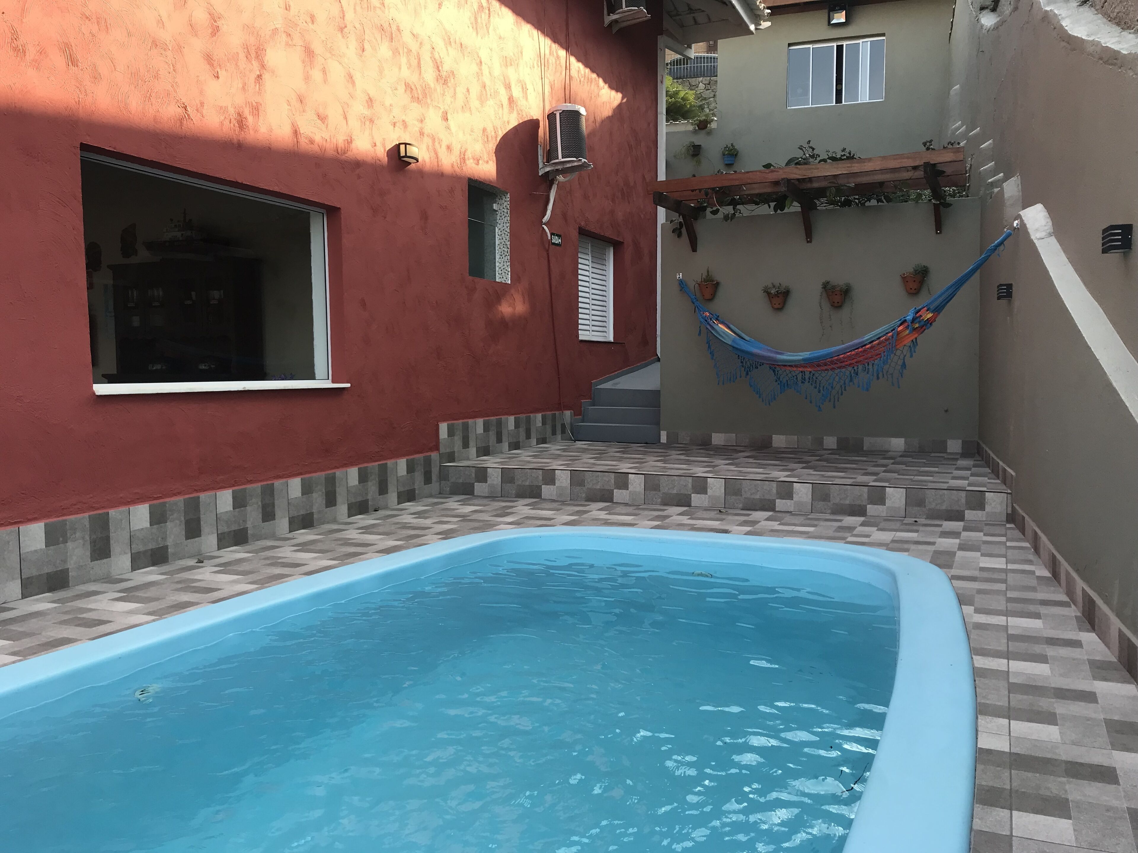 Piscina com chão revestido de azulejos, uma rede e um edifício com parede vermelha e janelas.