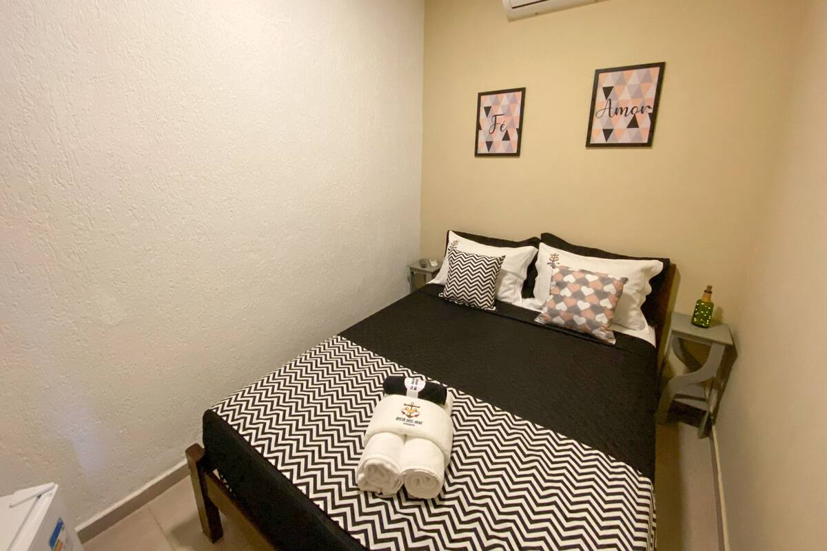 Cama bem arrumada com colcha em padrão ziguezague preto e branco, dois travesseiros e uma pequena garrafa em uma mesa de cabeceira.