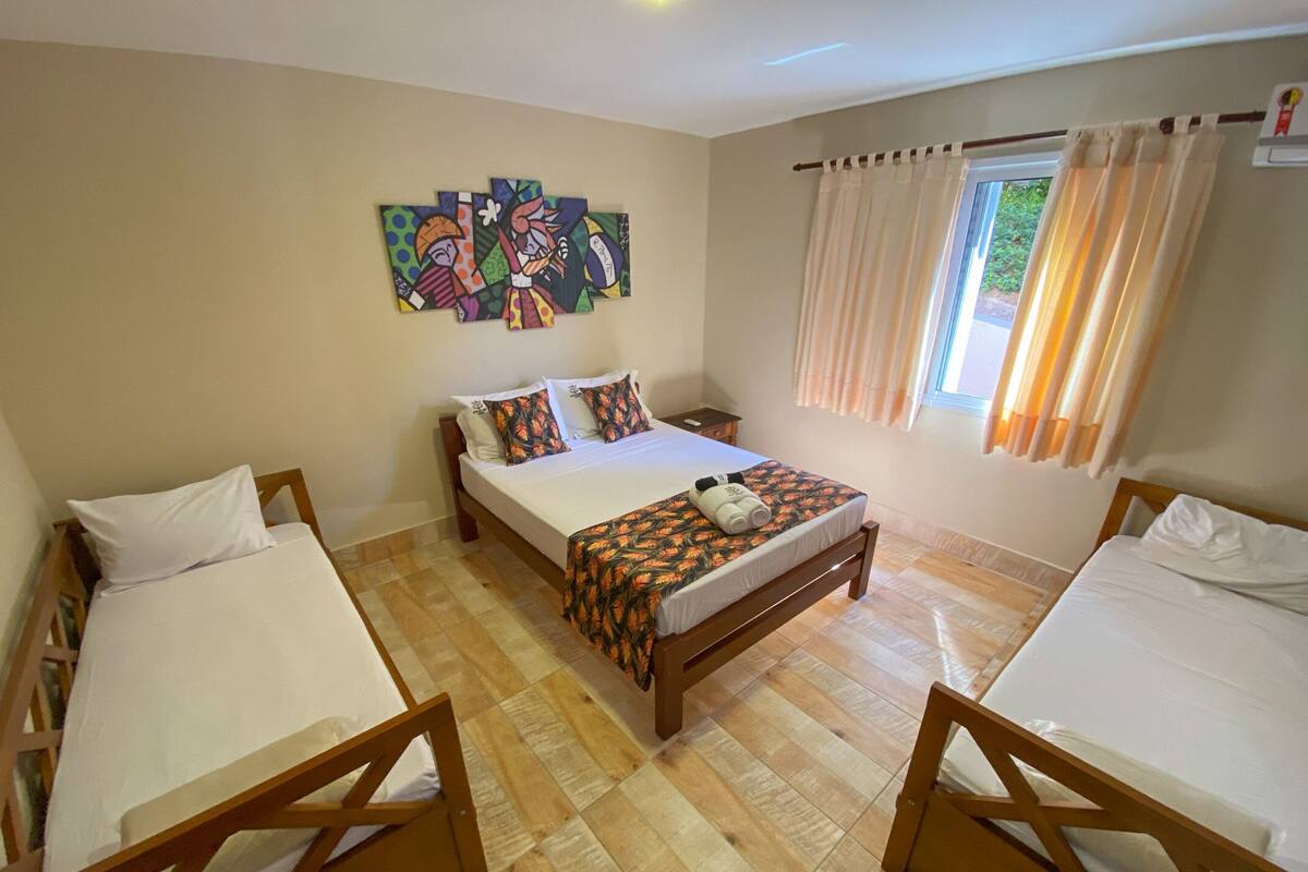 Quarto de hotel com uma cama grande, duas camas menores, piso de madeira, um quadro na parede e uma janela com cortinas.