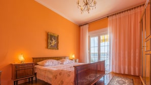 Quarto com cama de madeira, duas mesas de cabeceira com luminárias, uma janela grande com cortinas e um candelabro.