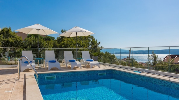 Piscina com espreguiçadeiras e guarda-sóis, cercada por guarda-corpo de vidro, com vista para um corpo d'água e paisagem distante.