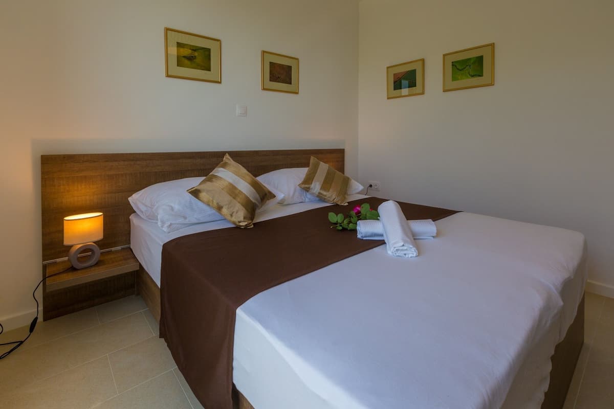 Cama bem arrumada com lençóis brancos, um cobre-leito marrom e duas almofadas. Há uma pequena mesa de cabeceira com um abajur e um cabo de energia. O quarto tem um design simples e limpo, com duas fotografias emolduradas na parede.