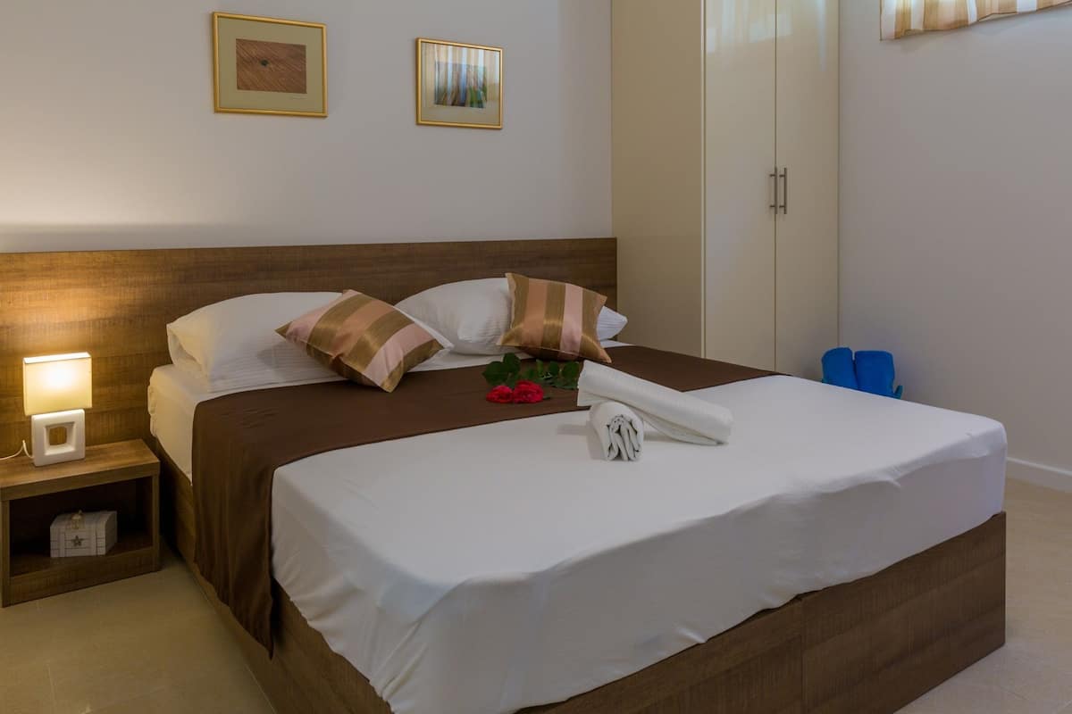 Cama bem arrumada com lençóis brancos, cabeceira de madeira e duas almofadas. Há uma mesa de cabeceira com uma luminária e uma pequena caixa.