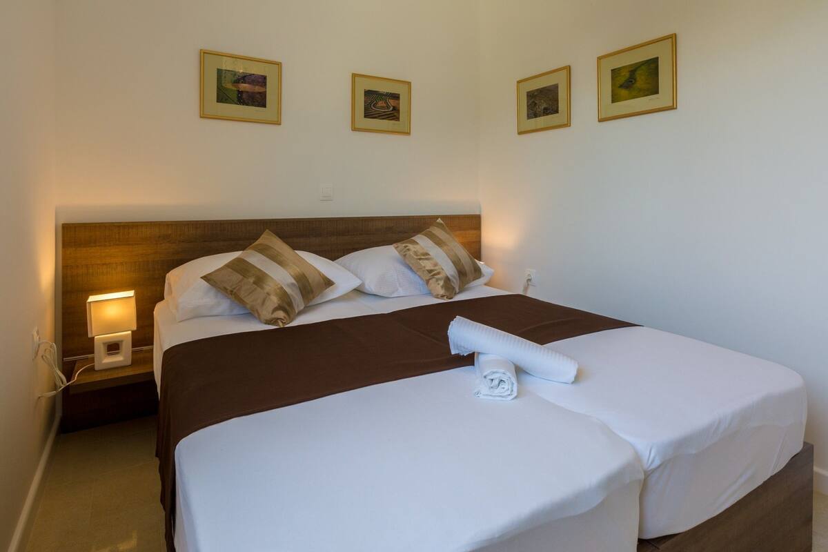 Quarto de hotel com duas camas, cabeceira de madeira e três quadros na parede.