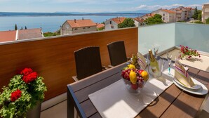 Uma varanda com uma mesa posta para dois, uma tigela de fruta, uma garrafa de champanhe e vista para o mar e edifícios.