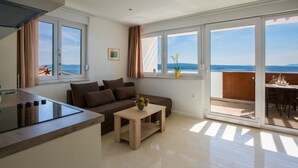 Sala de estar moderna com sofá, mesa de centro e área de cozinha com fogão. Janelas amplas oferecem vista para o mar e uma varanda com mesa de jantar.