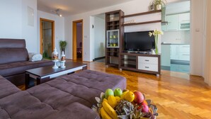 Uma sala de estar moderna com um sofá marrom, uma mesa de centro e uma tigela de frutas. Há uma TV em um suporte de madeira e uma área de cozinha visível através de uma porta aberta.