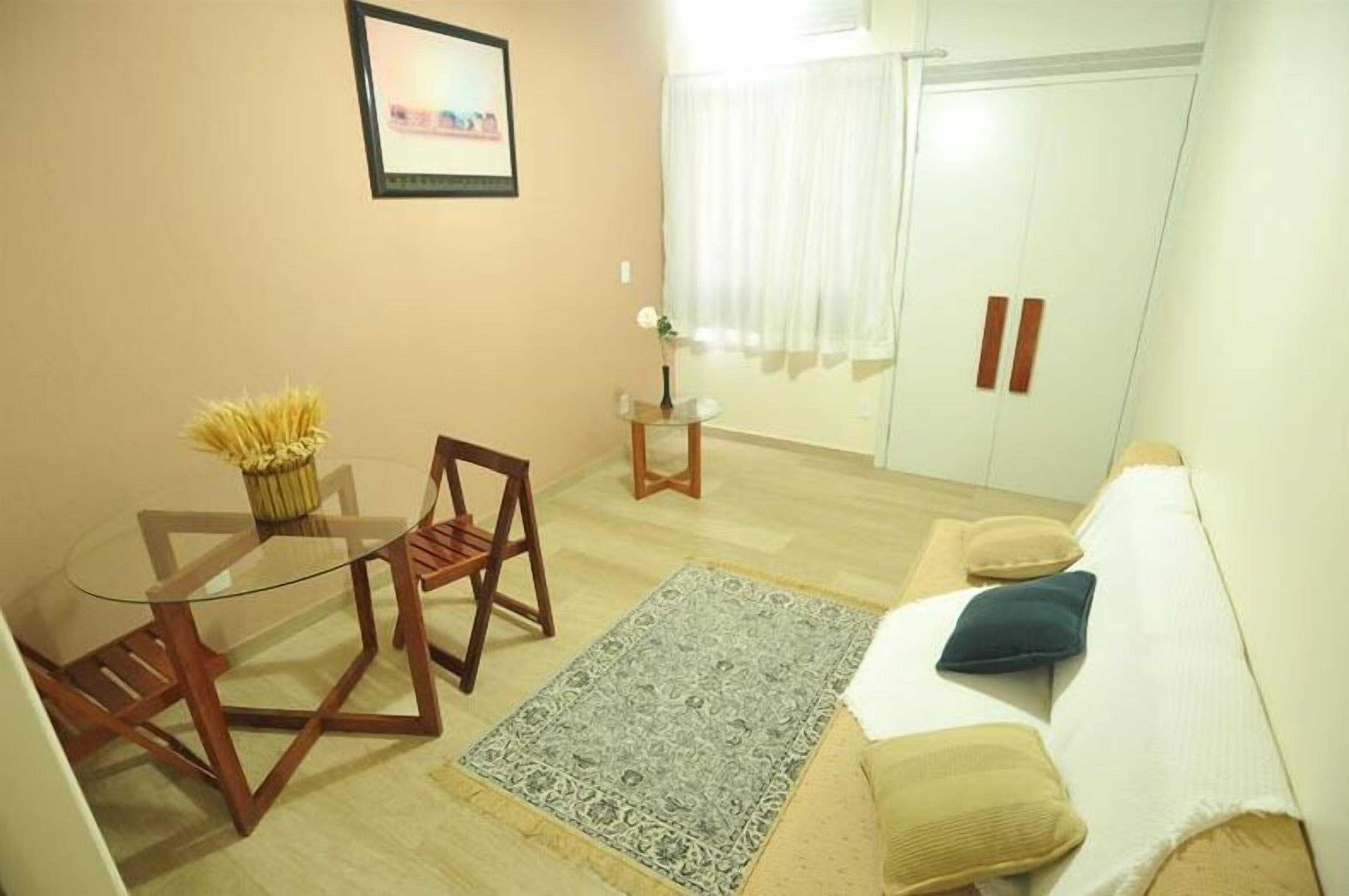 Quarto pequeno e bem iluminado, com mesa de jantar de vidro, duas cadeiras de madeira, um sofá com almofadas, tapete estampado, uma mesinha lateral com um vaso e um guarda-roupa branco com espelho.